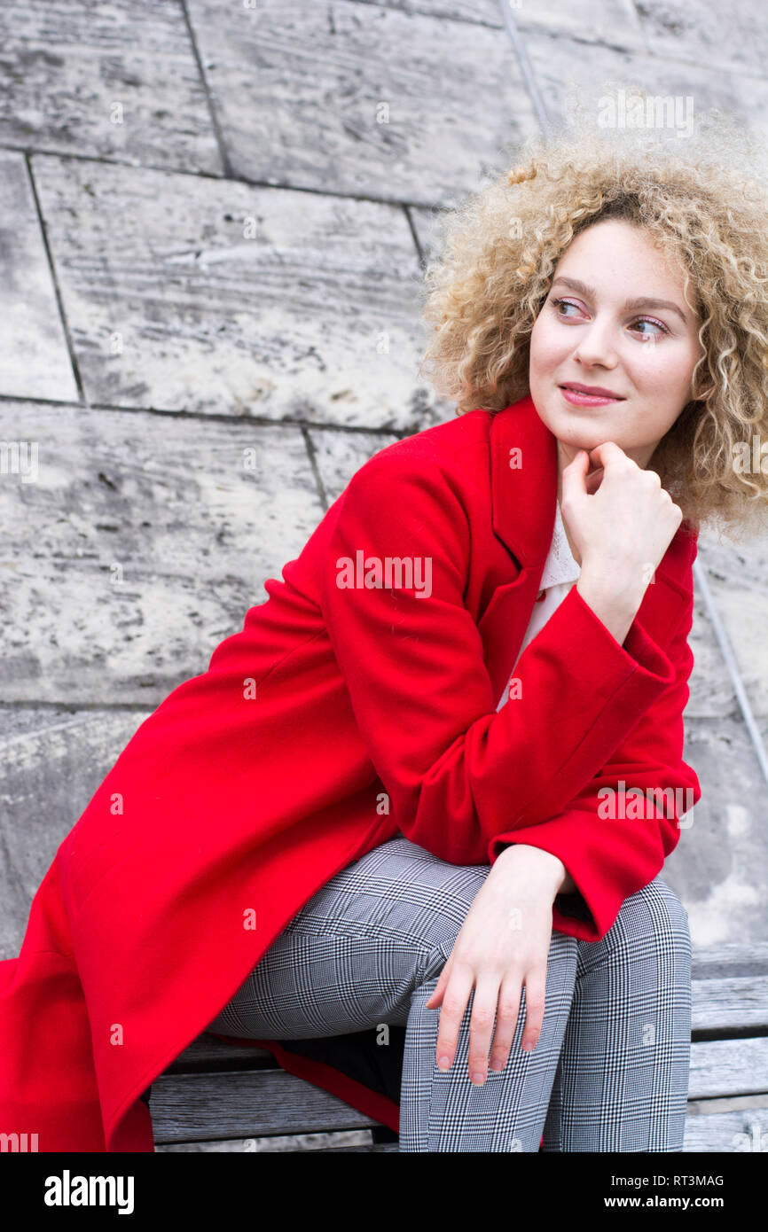 Portrait of smiling blond woman with ringlets wearing red coat watching ...