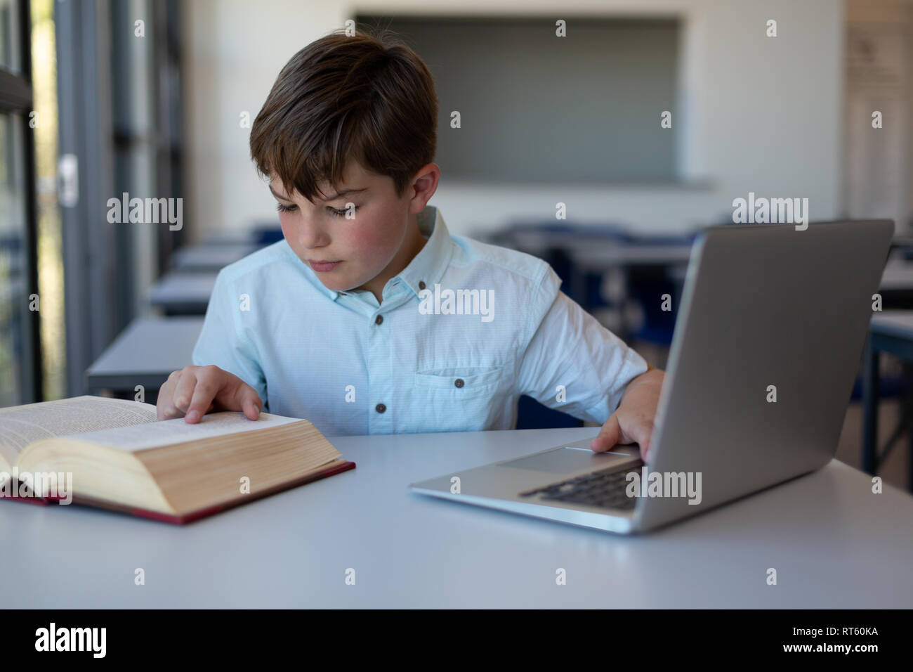 Schoolboy reading a book and using laptop in a classroom Stock Photo ...