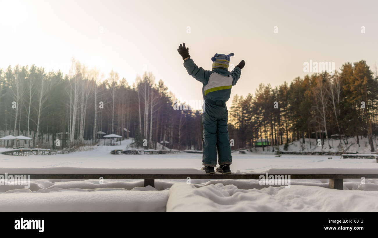 Boy at the frozen lake at winter enjoys sunlight, raising his hands up ...