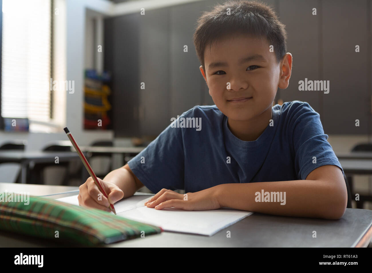 Schoolboy looking at camera while writing on a book at desk in ...
