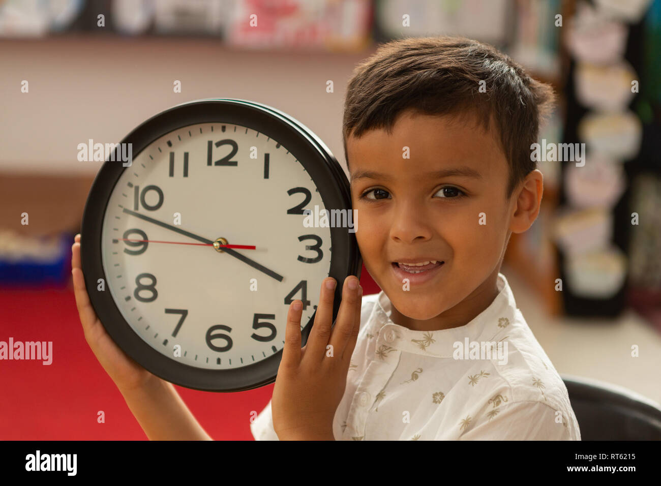 Smiling schoolboy with wall clock looking at camera in a classroom ...