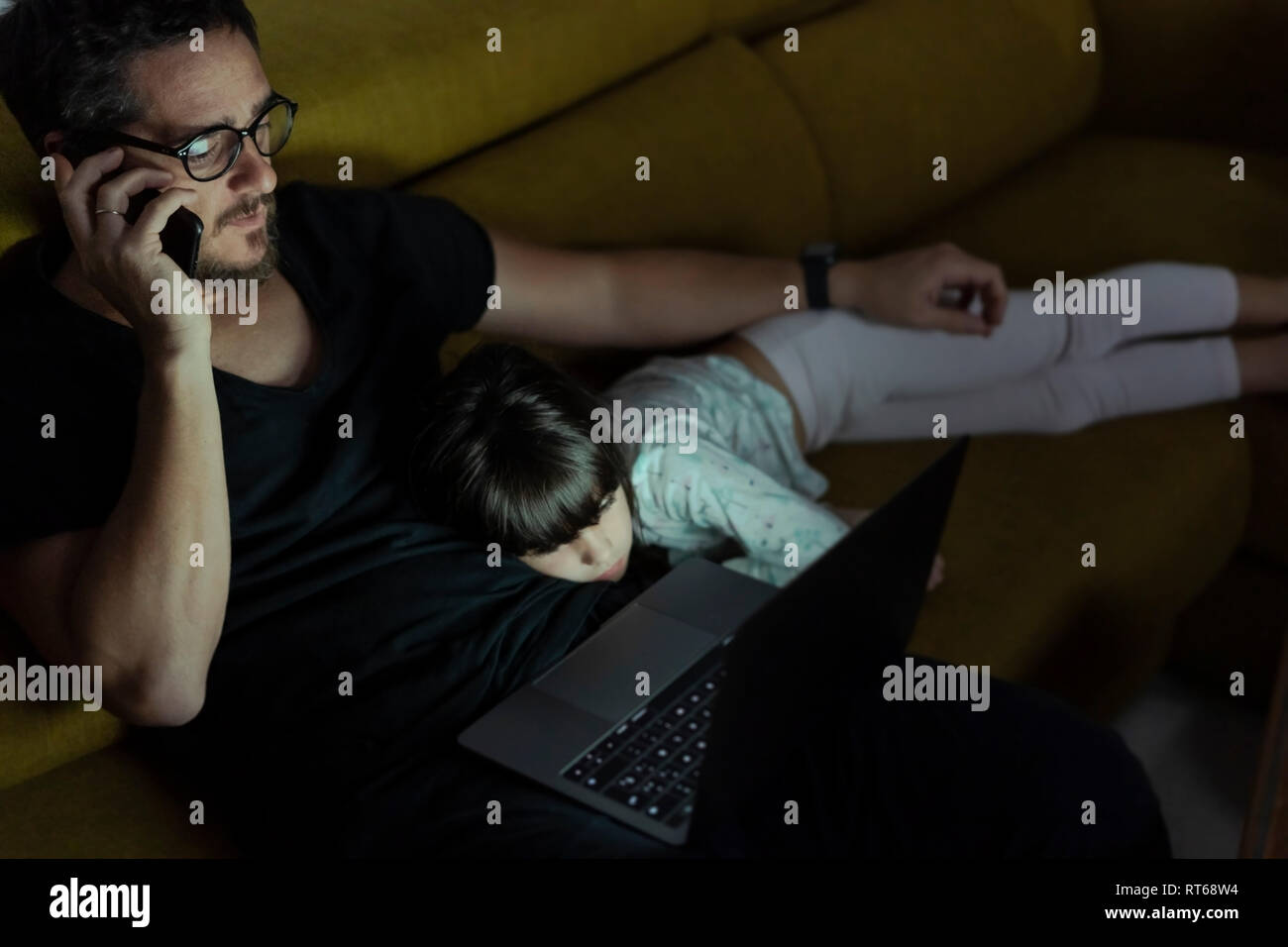 Father talking on cell phone on couch at night with daughter lying next ...