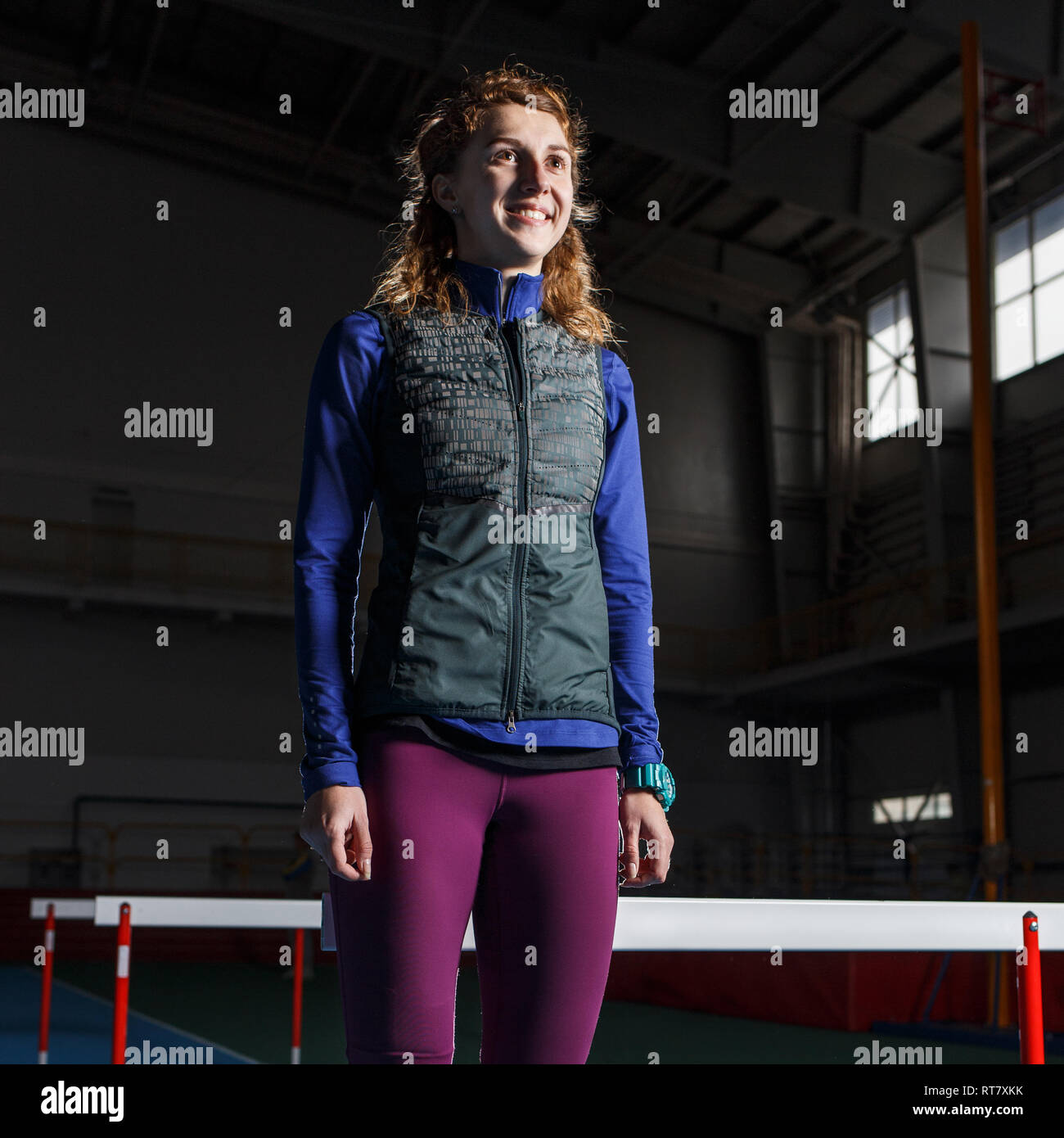 Young smiling female athlete standing near hurdle on sprint lane ...