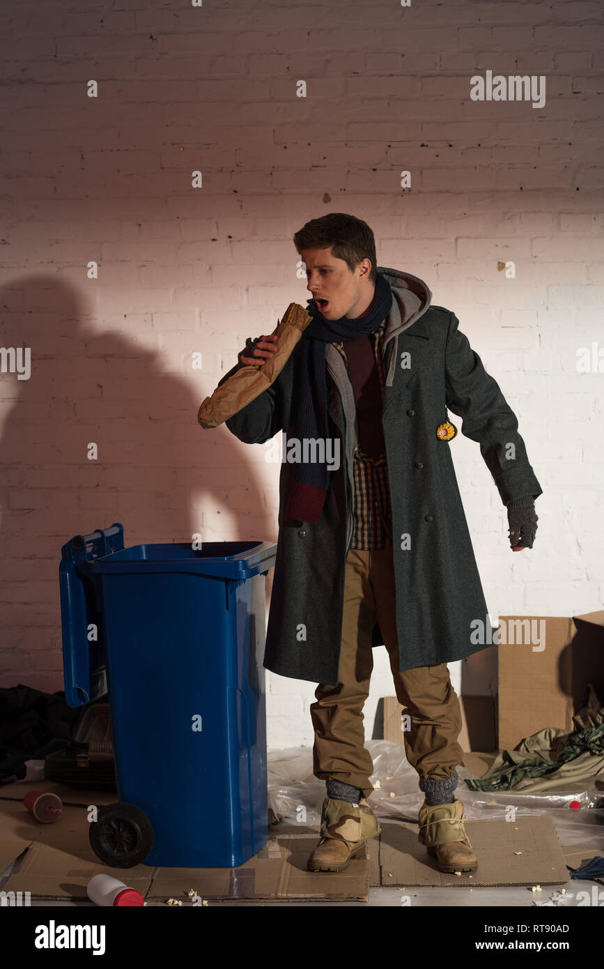 hungry homeless man eating bread baguette while standing by trash ...