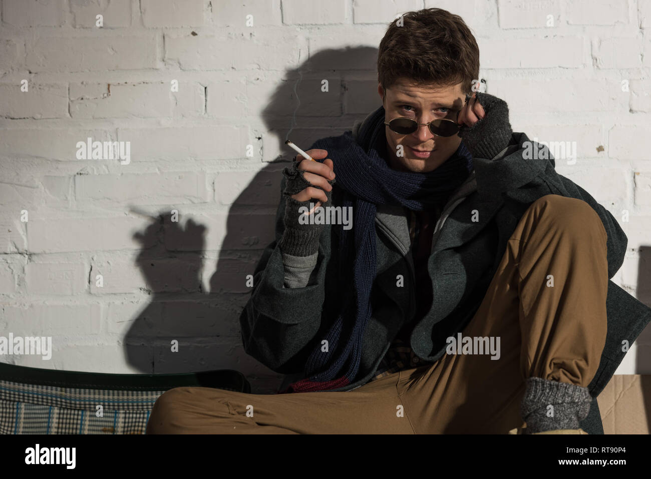 homeless man in dark glasses smoking while sitting by white brick wall ...