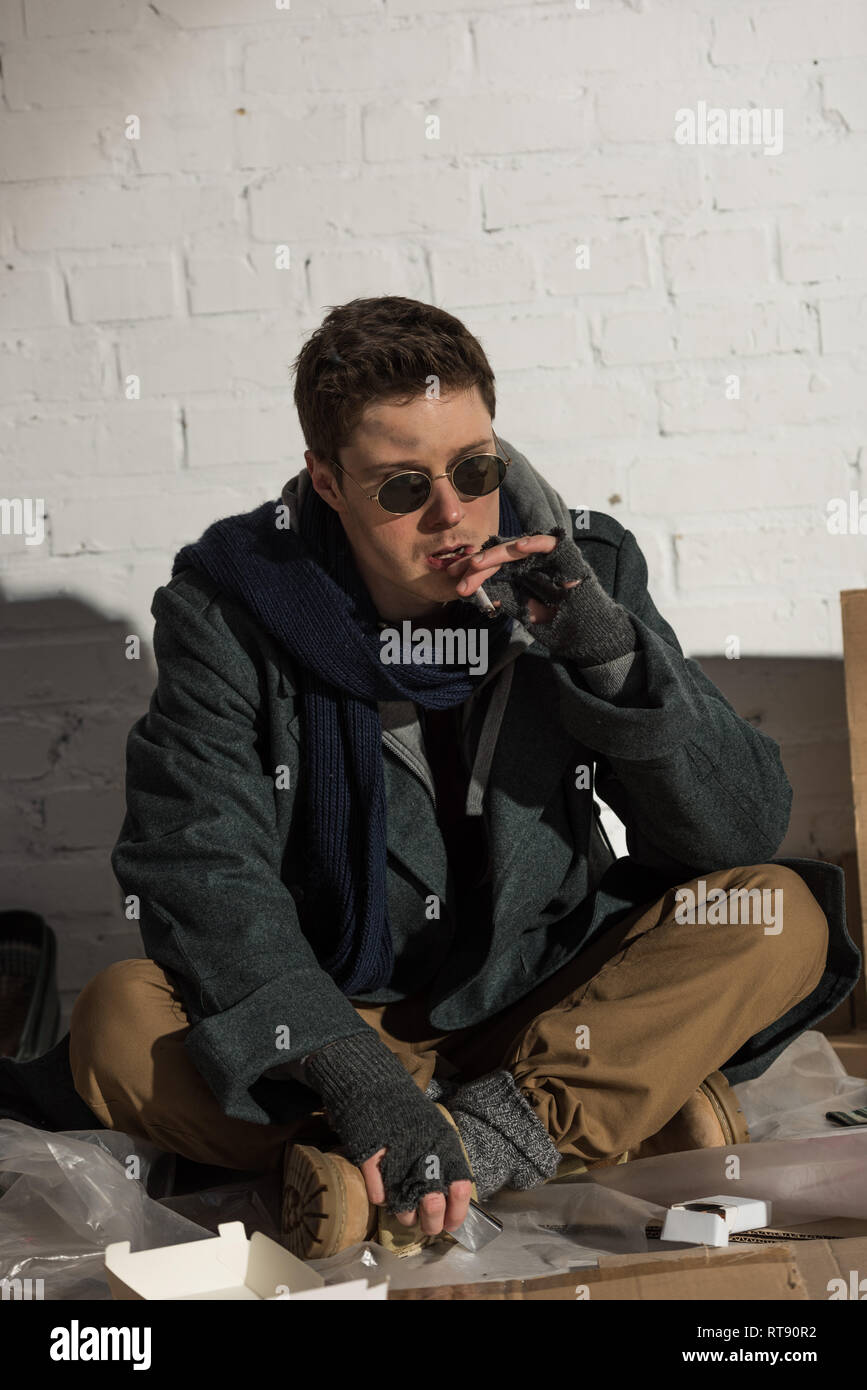 homeless man smoking cigarette while sitting by white brick wall Stock ...