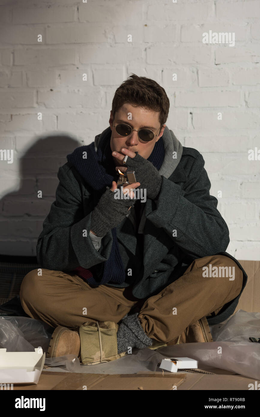 homeless man lighting cigarette while sitting by brick wall Stock Photo ...