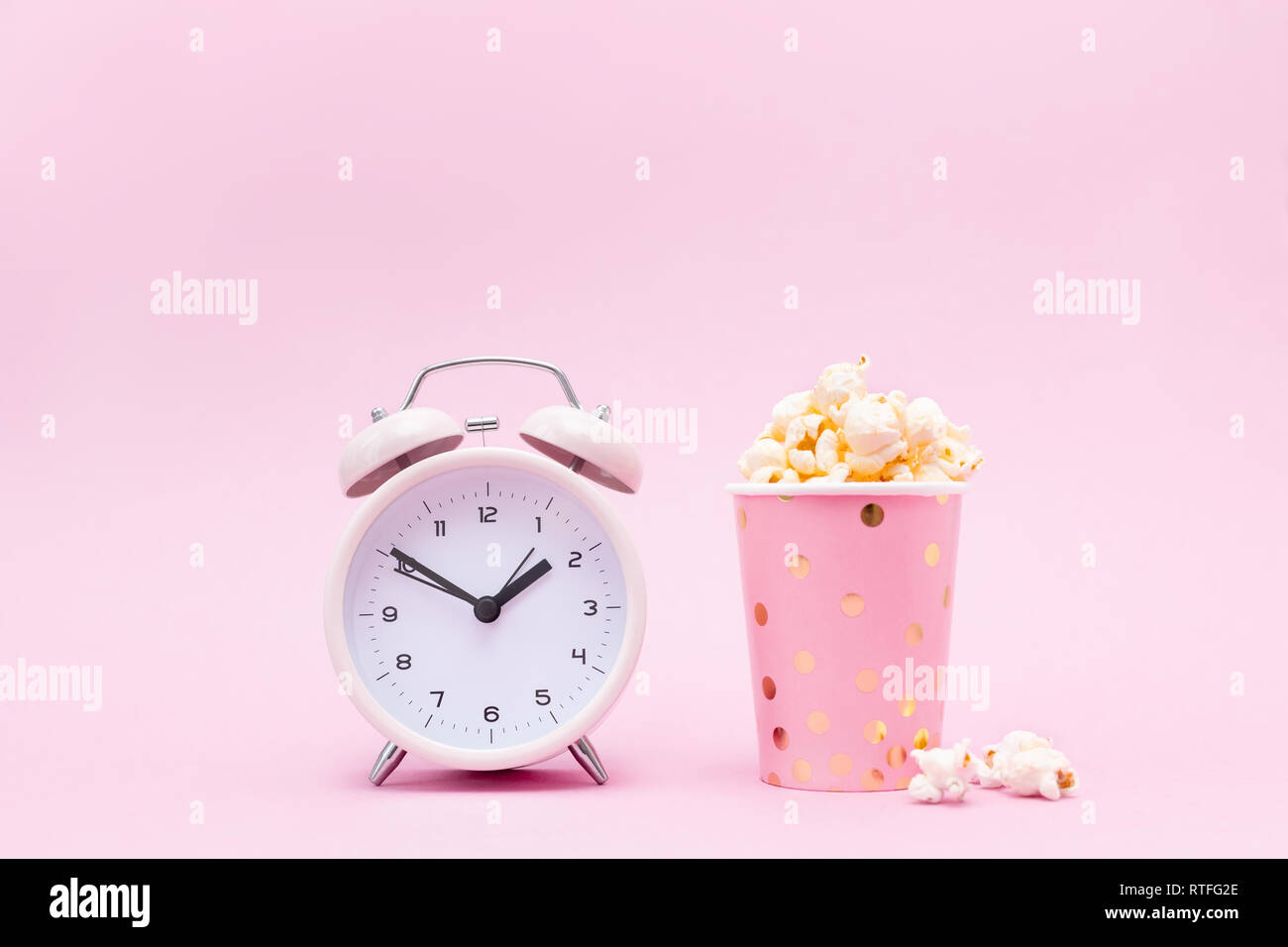 Popcorn in a bright glass and alarm clock on a pink background. Time ...