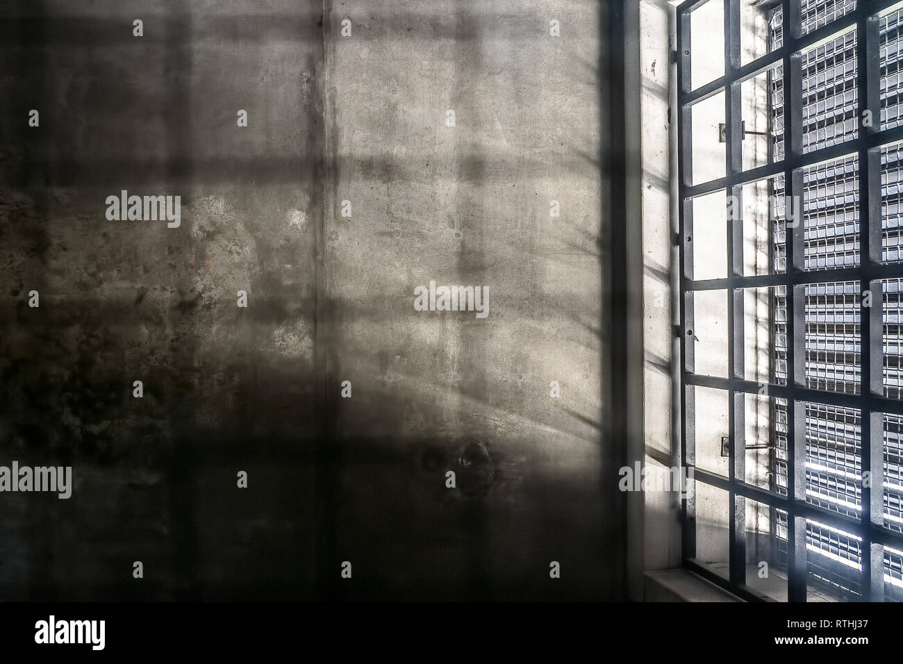 The very sober interior of a prison cell: barred windows with little light coming in and bare ...