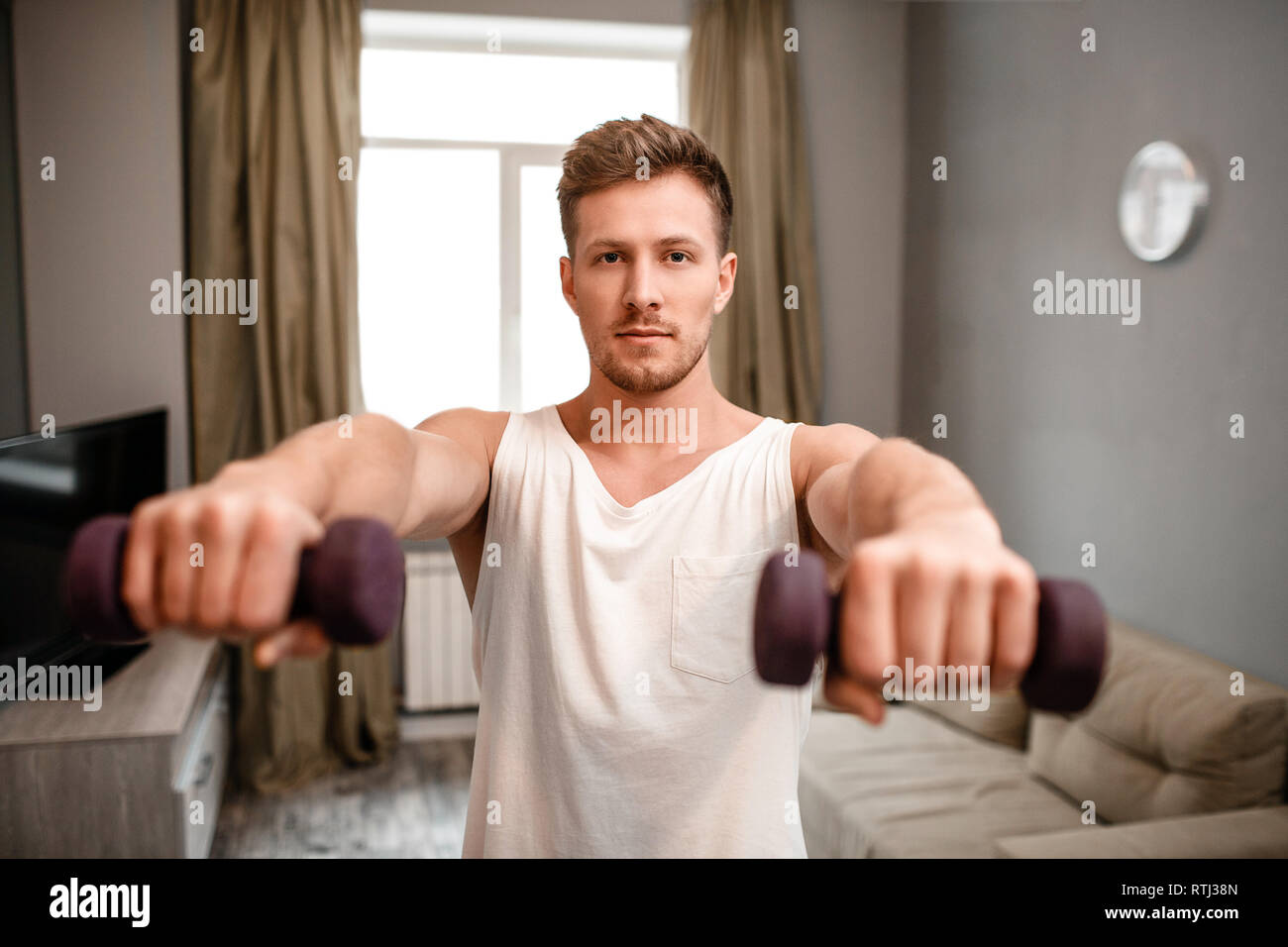 Young well-built man go in for sports in apartment. He hold dumbbells ...