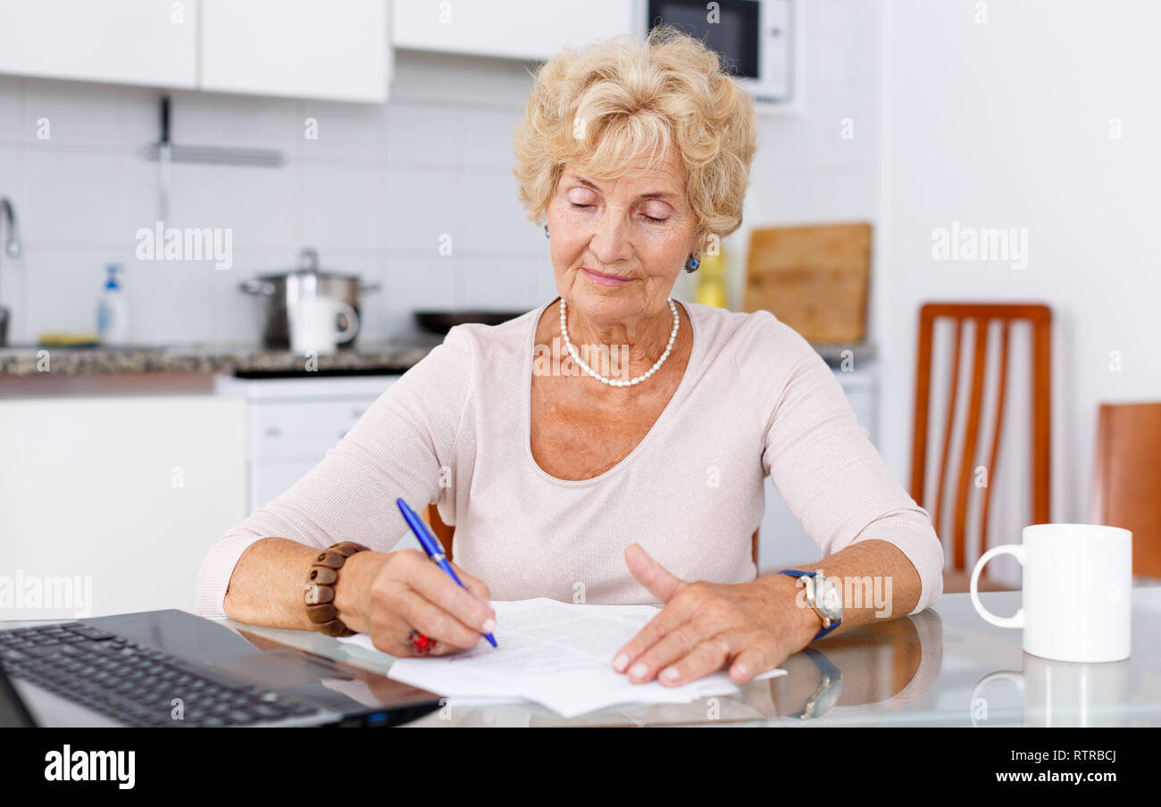 Senior woman sitting and filling up documents at kitchen table Stock ...