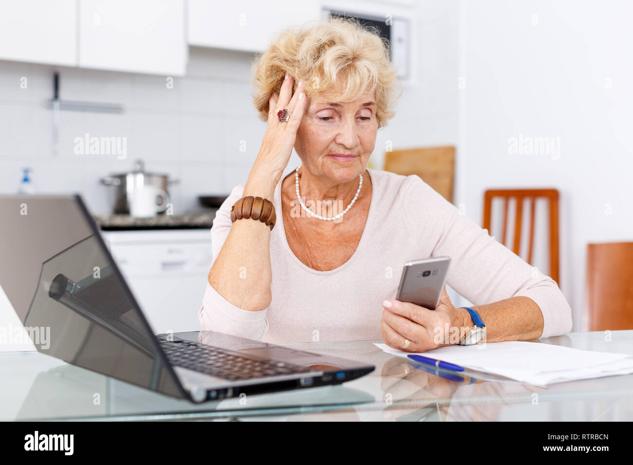 Confused elderly woman using laptop and smartphone at kitchen table ...