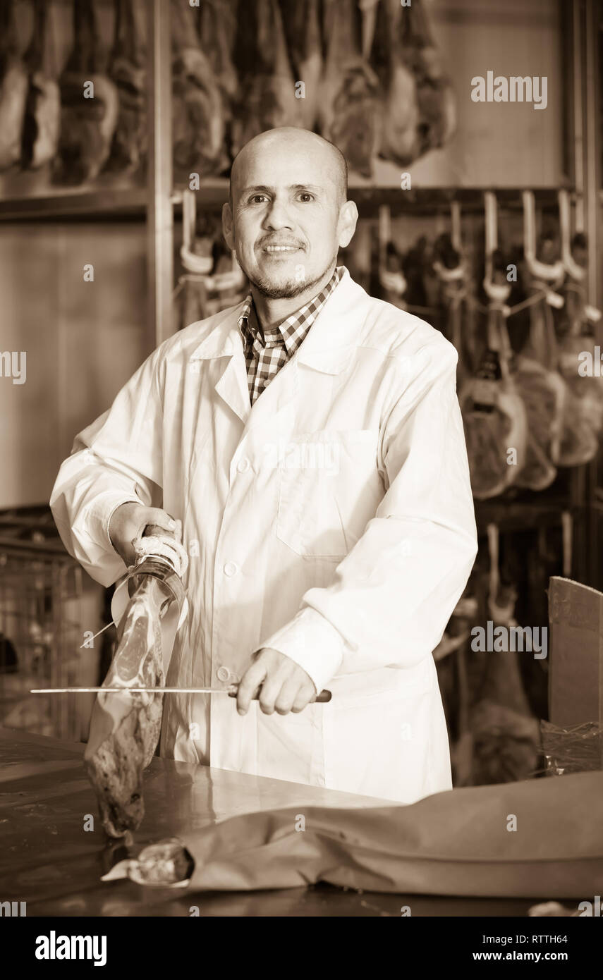 Mature butcher slicing jamon and smiling in delicatessen store Stock ...