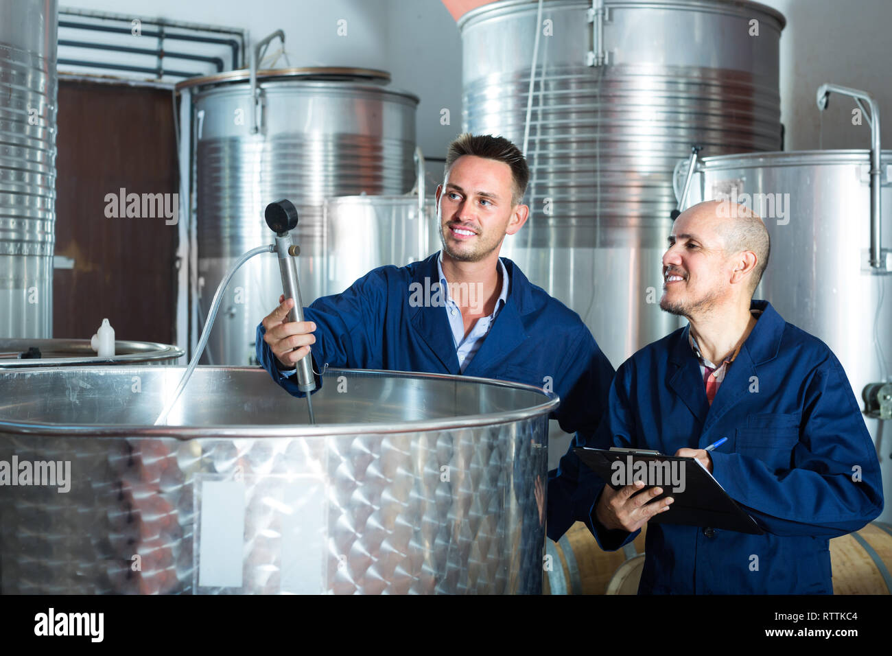 Two cheerful male winery workers in uniform working and taking notes in ...