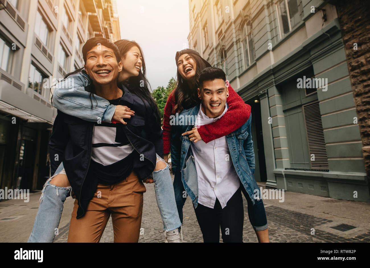 Two young asian men carrying women friends on their back on city street ...