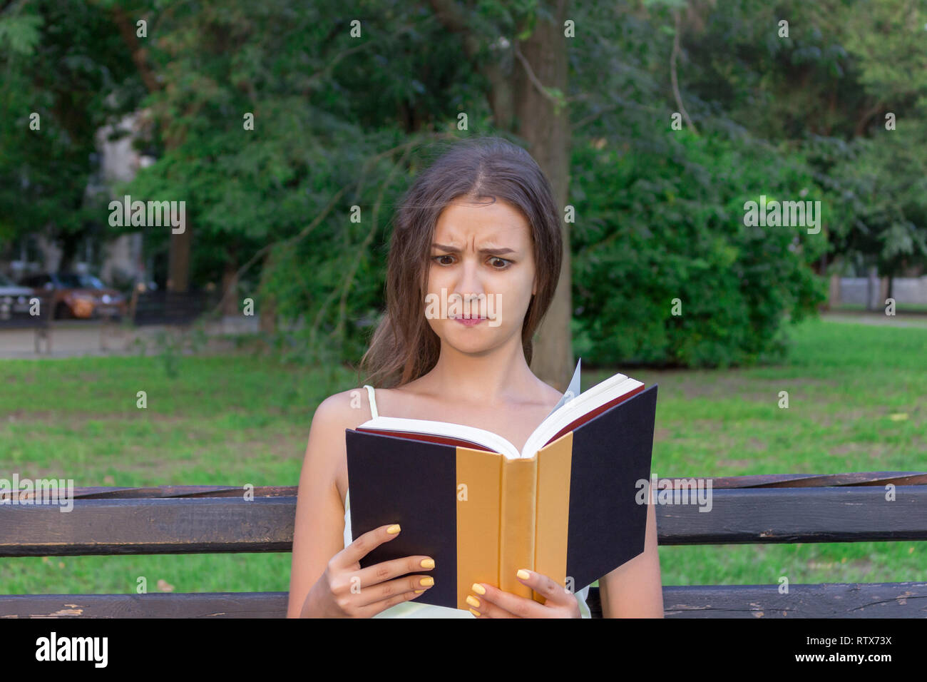 Confused and displeased girl is reading a book on the bench in the park ...