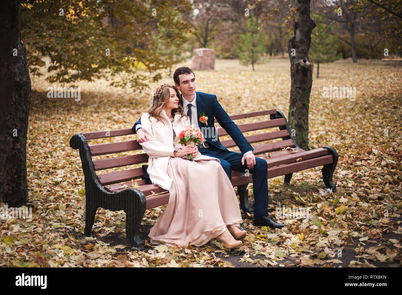 The beautiful bride and handsome groom sitting on a bench in the autumn ...