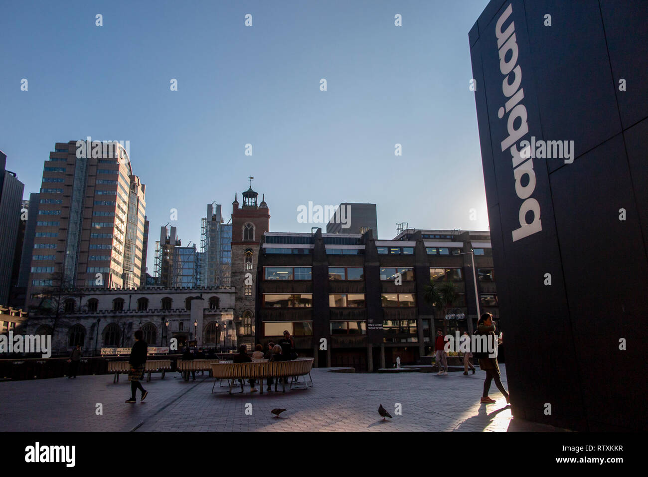 A large Barbican sign with people walking past with long shadows from ...