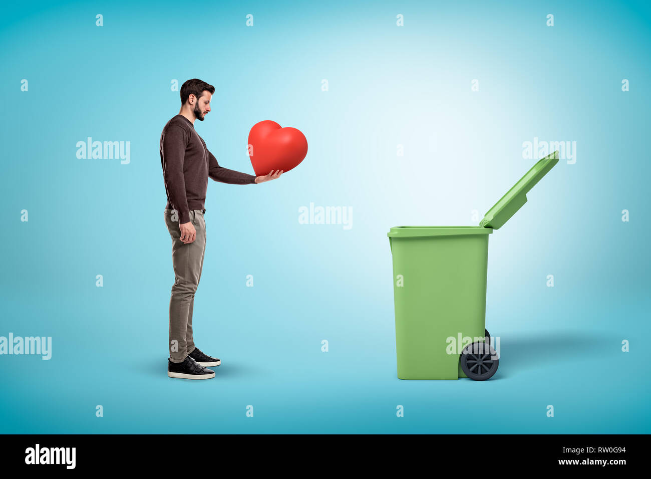 Man throwing red heart into open garbage bin on blue background Stock ...