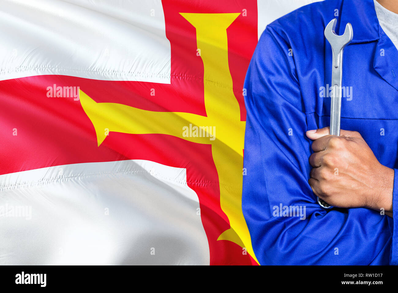 Mechanic in blue uniform is holding wrench against waving Guernsey flag ...