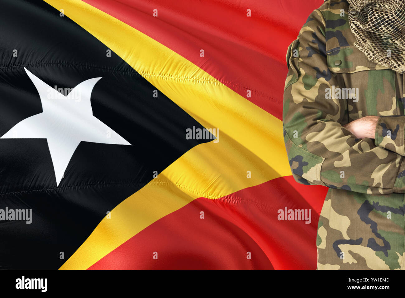 Crossed arms Timorese soldier with national waving flag on background ...