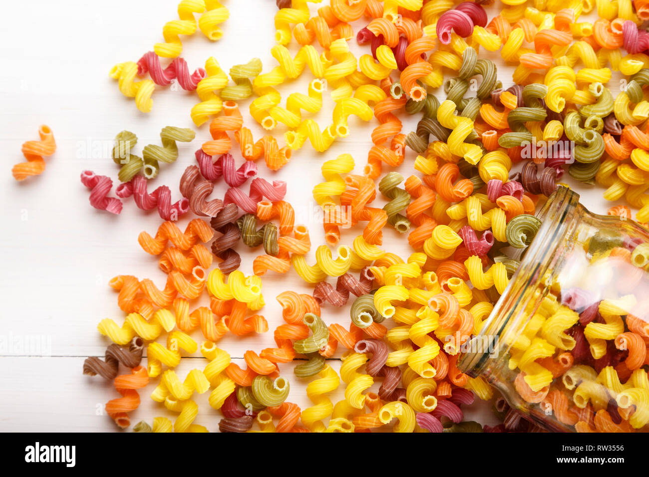 uncooked multi-colored pasta poured from a jar on a white table. space ...