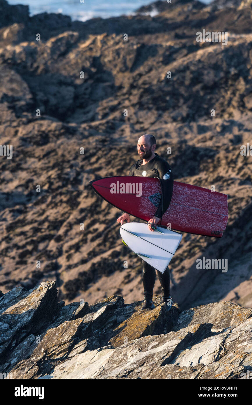 A disconsolate surfer standing on rocks carrying his snapped surfboard ...
