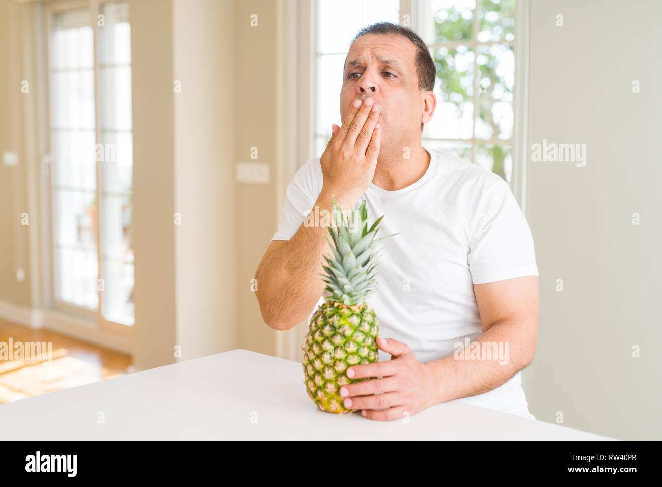 Middle age man eating fresh tropical pineapple at home cover mouth with ...