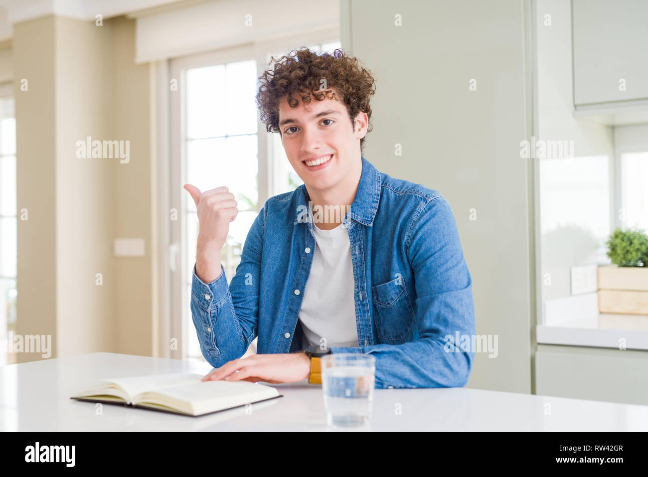 Young man reading a book at home pointing and showing with thumb up to ...