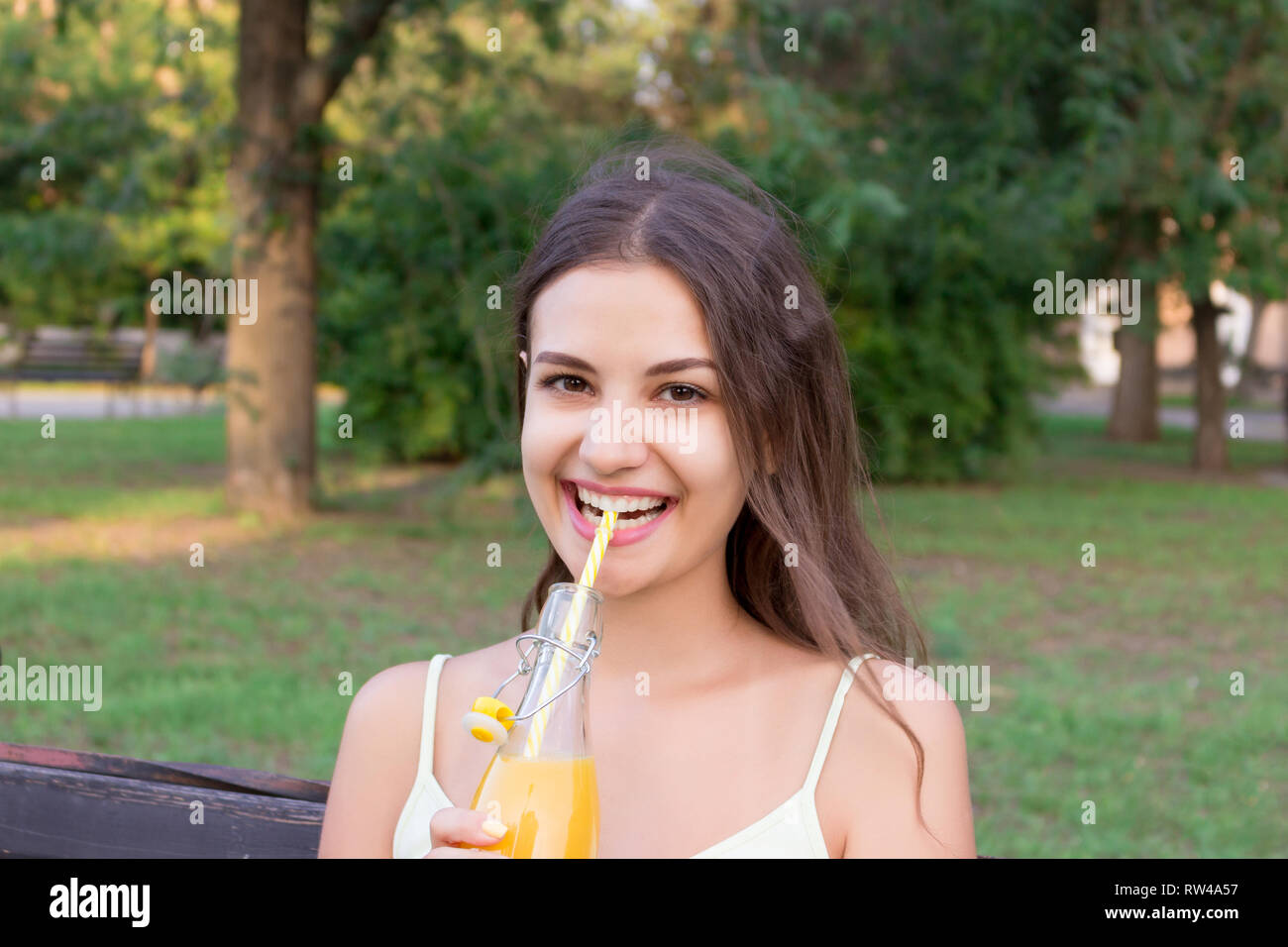 Young pretty girl is having rest on the bench in the park and drinking ...