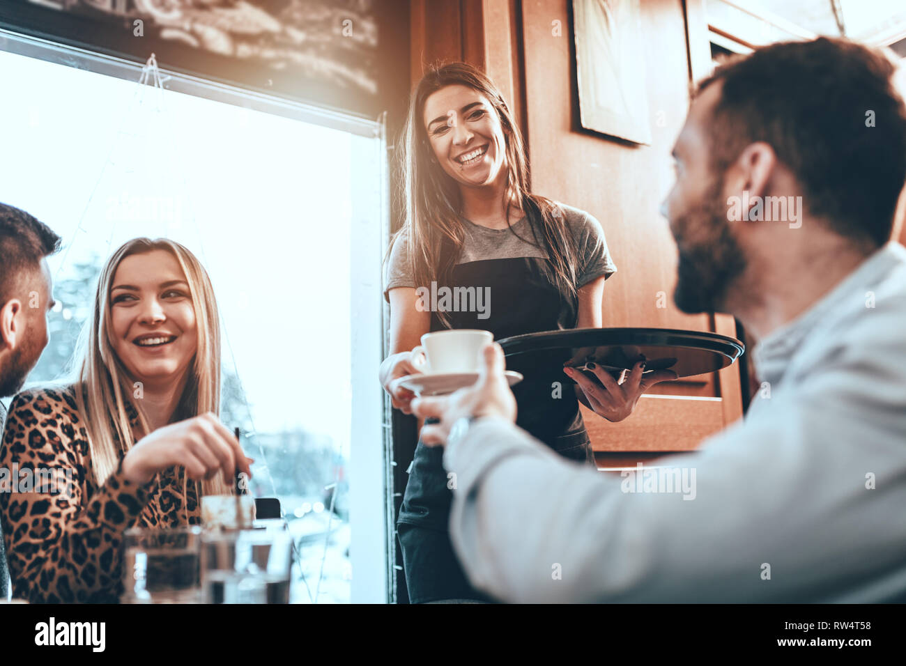 A young beautiful waitress is serving a group of young smiling friends ...