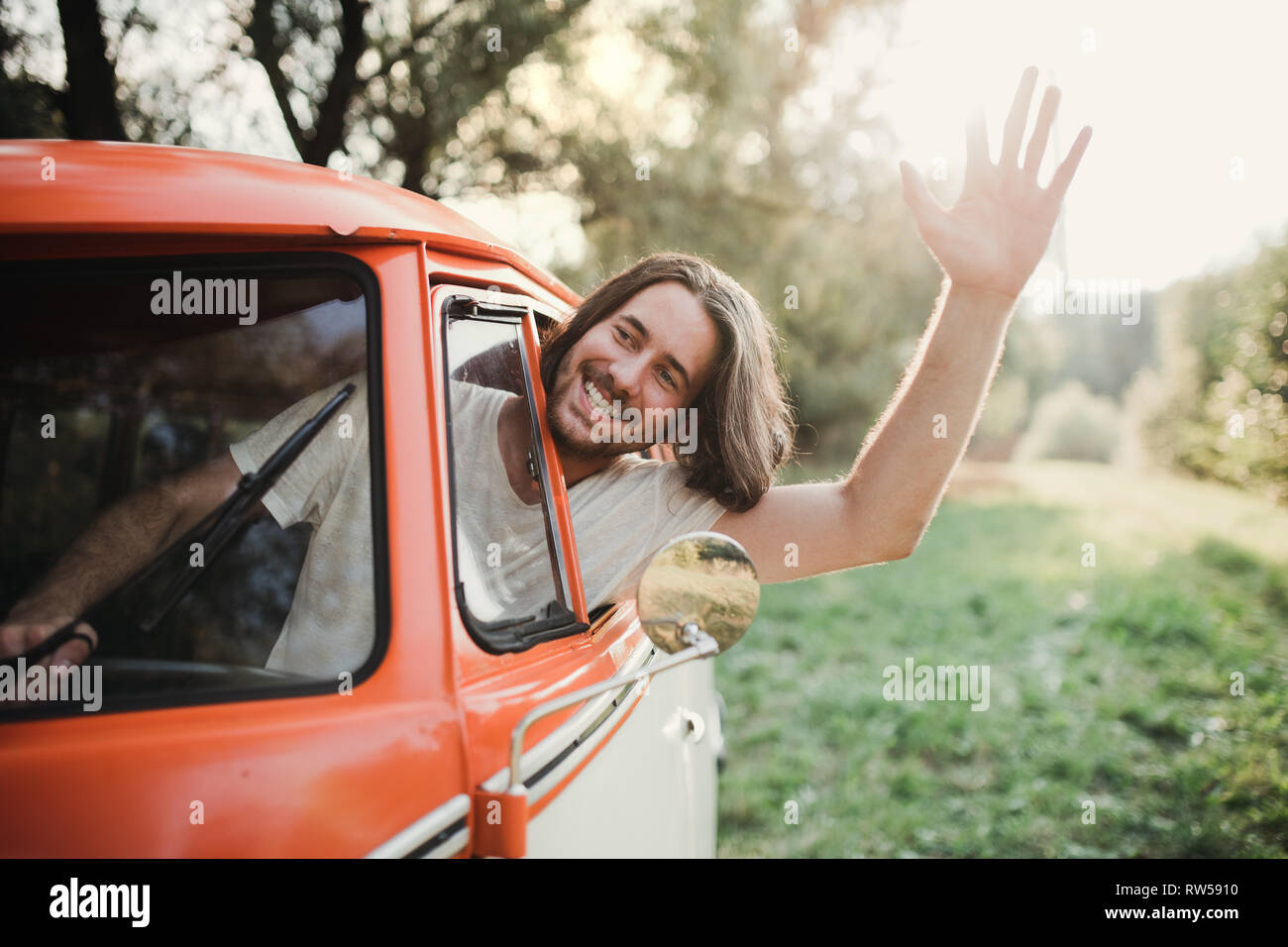 A young man driver in a car on a roadtrip through countryside, waving ...