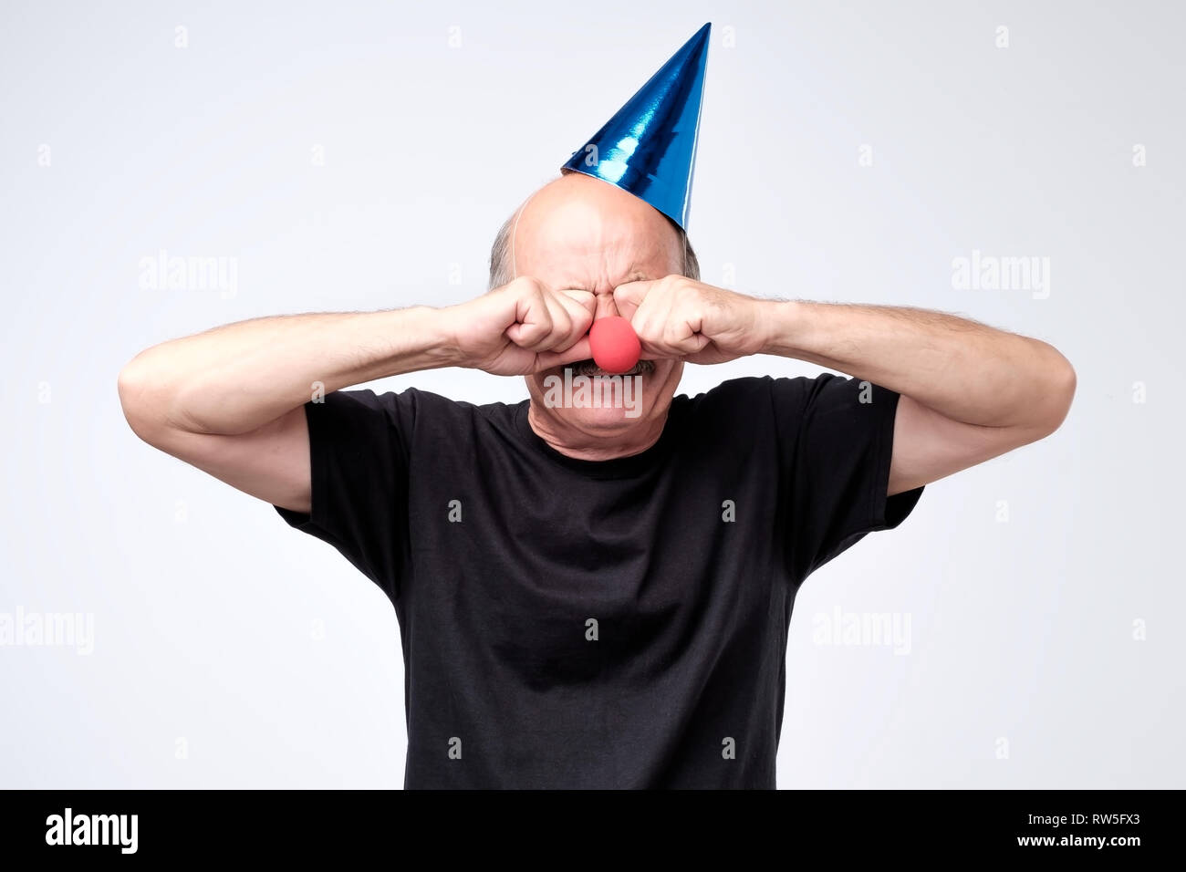 Dissatisfied disconcerted senior man in birthday cap crying and wiping ...