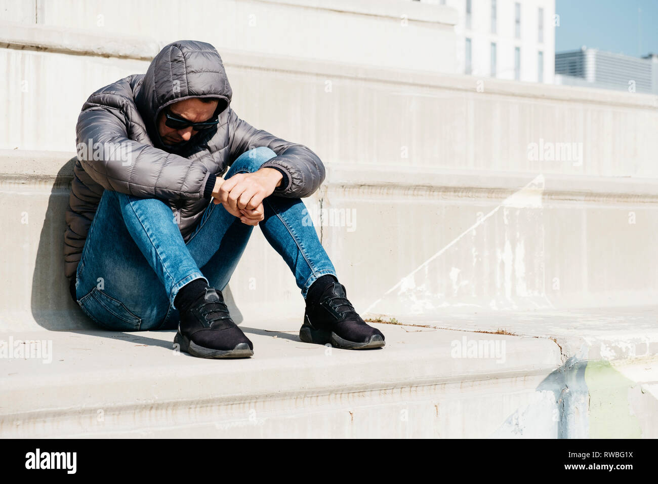 a young caucasian man, wearing jeans and a gray hooded jacket, curled ...