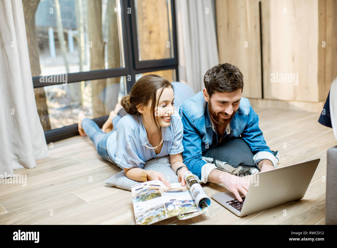 Happy couple lying on the floor with laptop and magazines, relaxing at ...