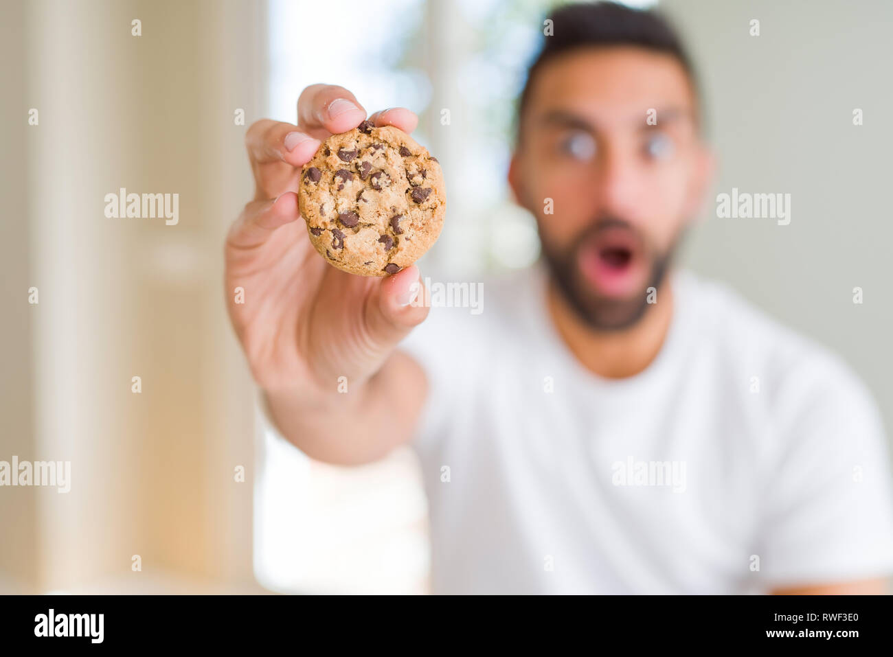 Handsome hispanic man eating chocolate chips cookies scared in shock ...