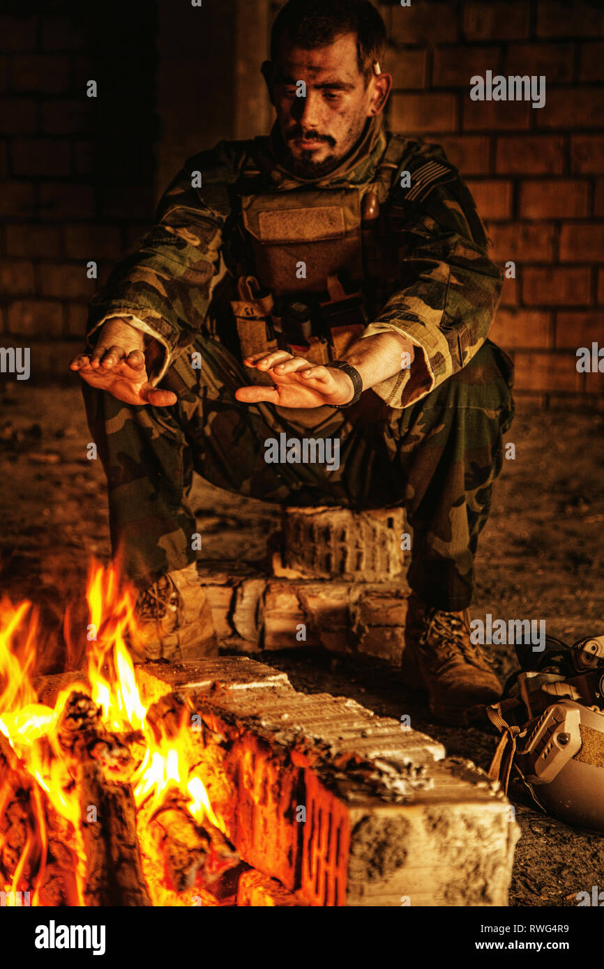 Special forces soldier warming his hands over a fire in a ruined ...