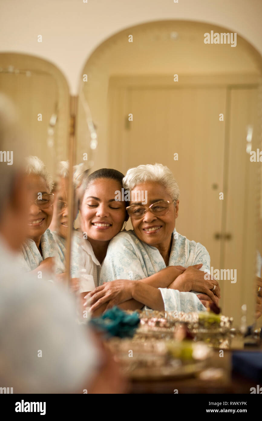 Daughter hugs her mother as they sit at a bedroom dresser with a mirror ...