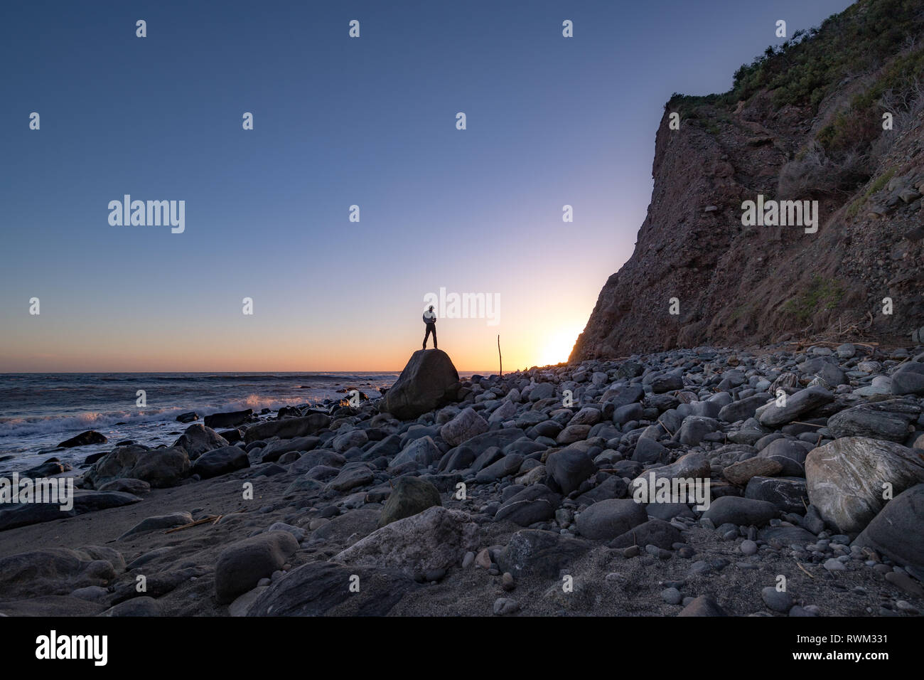 Beachside view of a man posed on a rock near a tall cliff as the sun ...