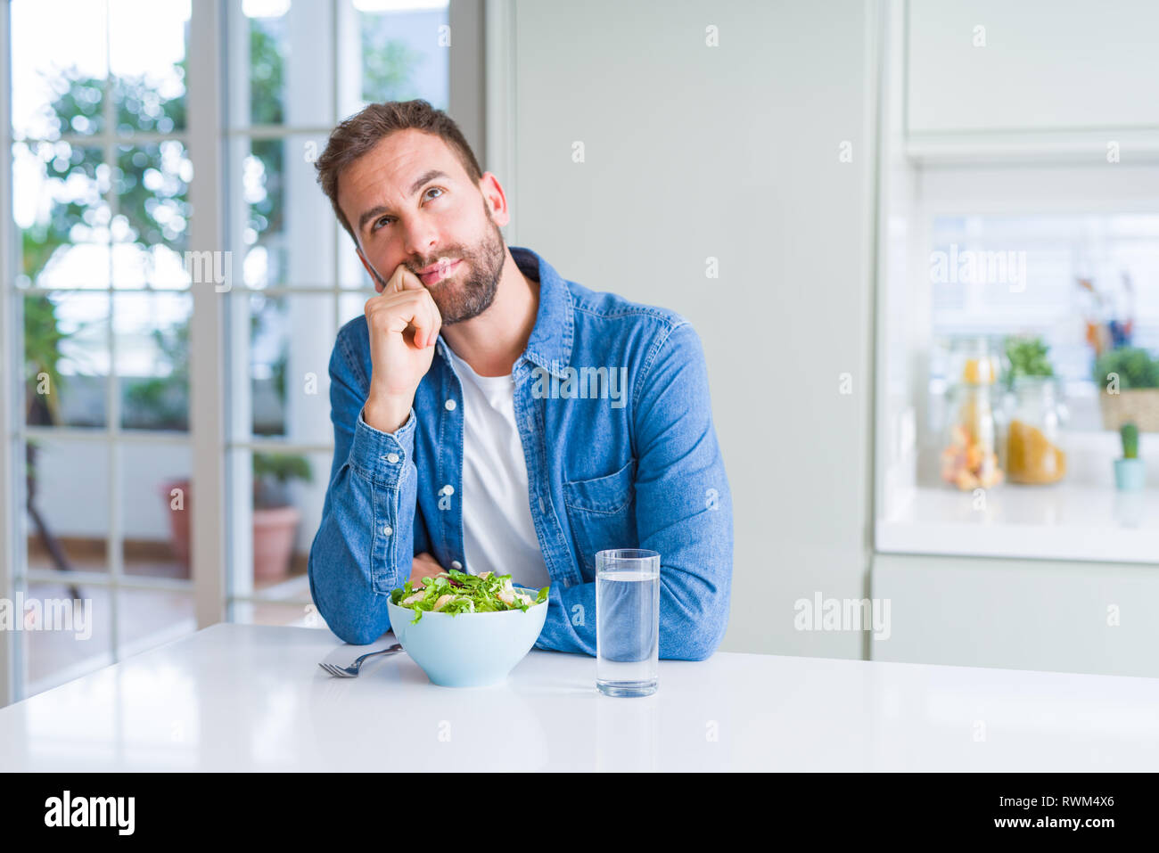Handsome man eating fresh healthy salad with hand on chin thinking ...