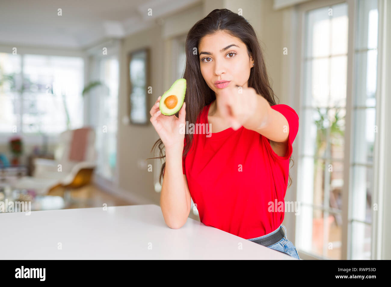 Young woman eating healthy avocado pointing with finger to the camera ...