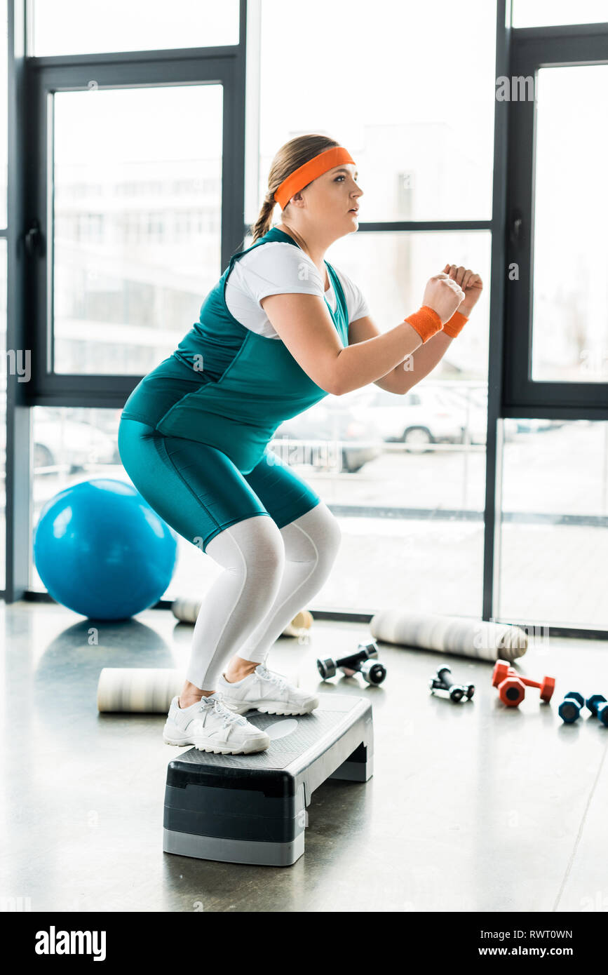 plus size woman squatting on step platform near fitness mats and ...