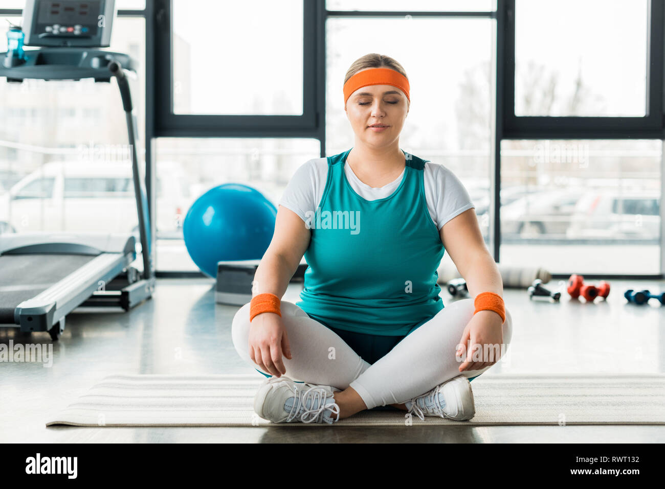 plus size woman sitting on fitness mat near sport equipment with ...