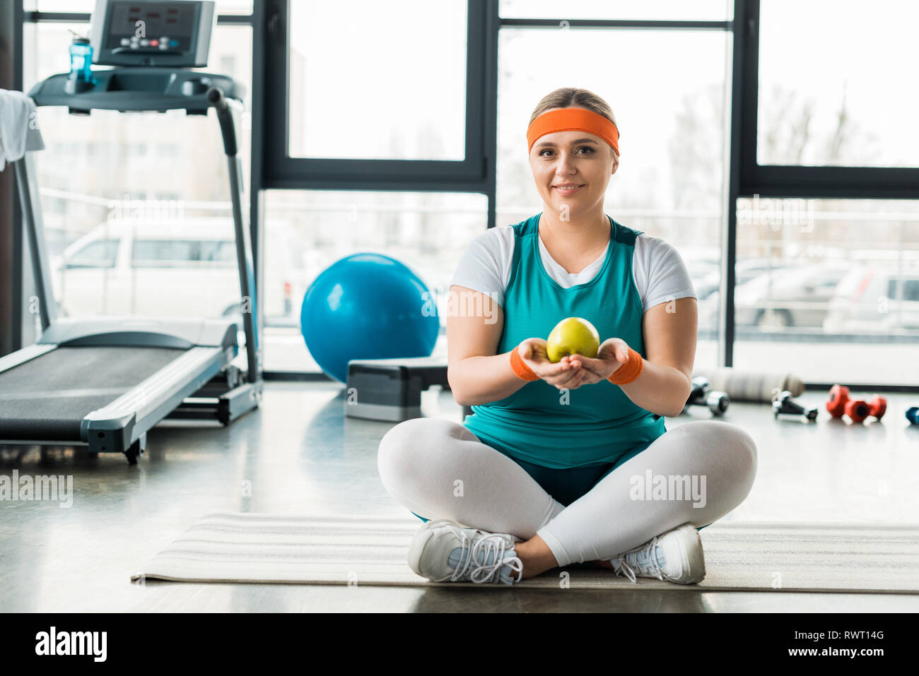 plus size woman sitting on fitness mat near sport equipment with ...