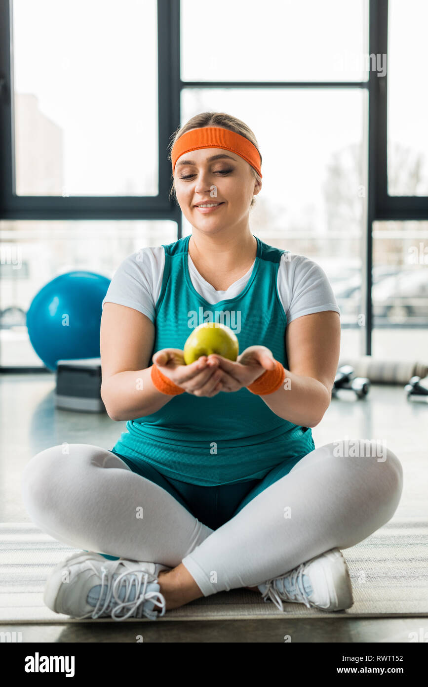 happy plus size woman sitting on fitness mat with crossed legs and ...