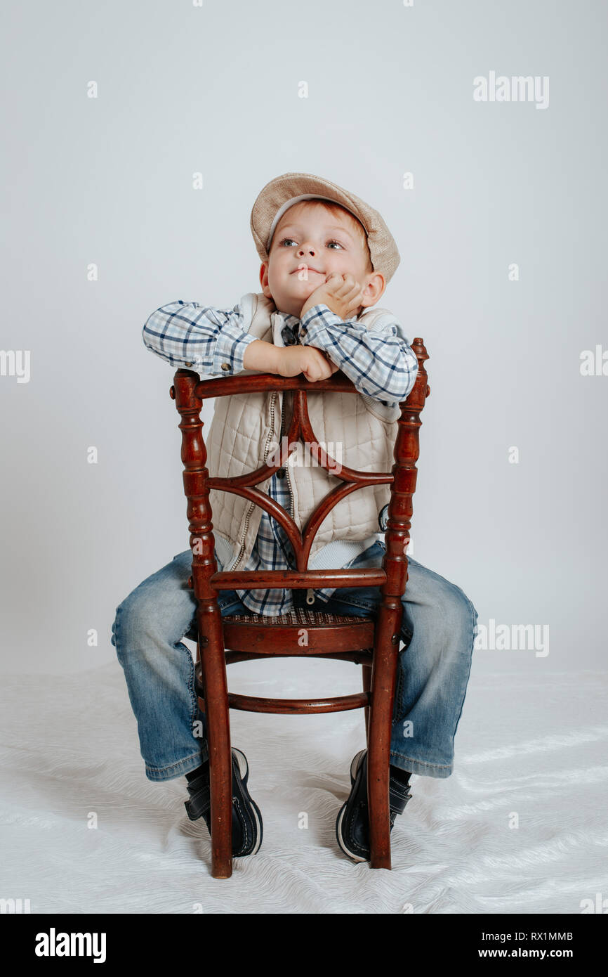 A little boy in a cap sits on a chair and smiles. On a white background ...