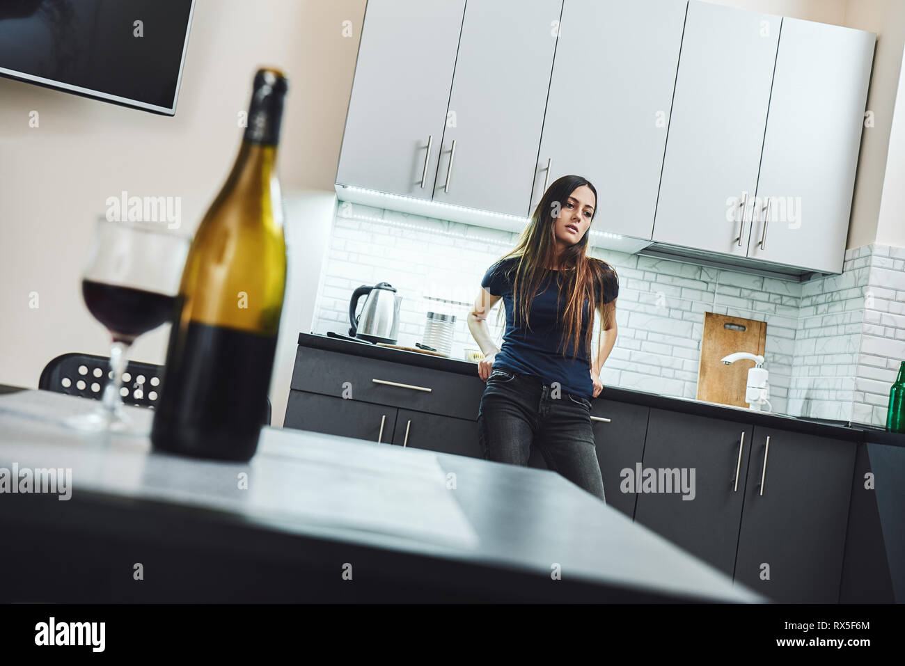 Bottle and glass of red wine in kitchen. Woman standing, looking ...