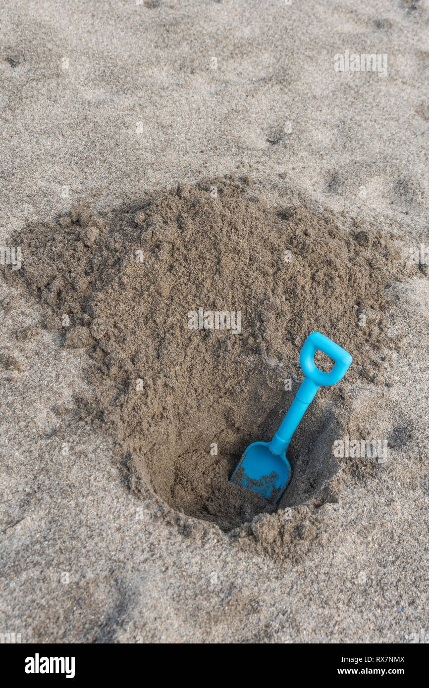 Seaside hole dug into sand, with kids blue spade. Metaphor 'dig a hole ...