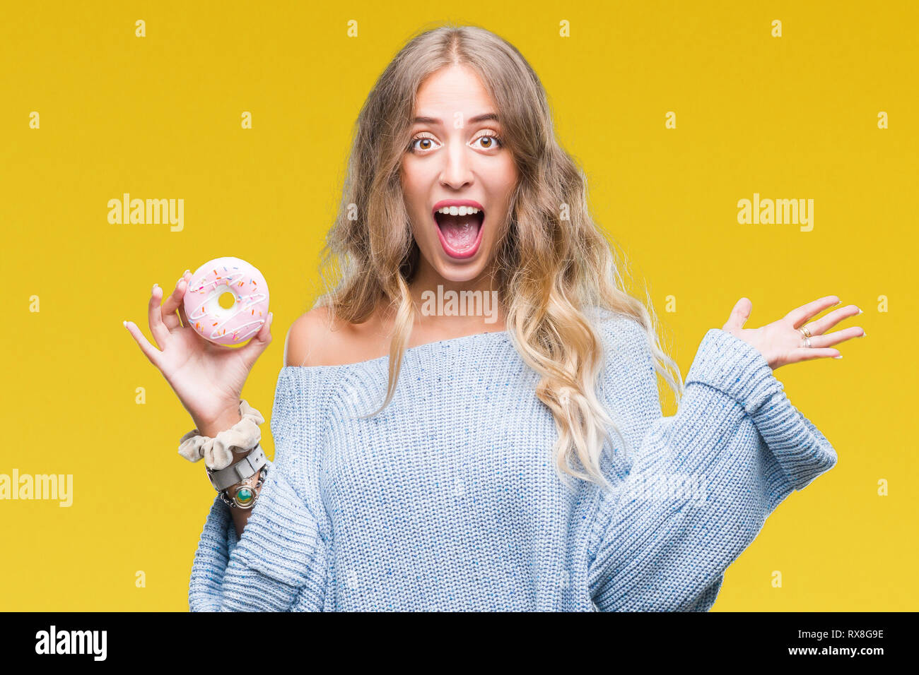 Beautiful young blonde woman eating pink donut over isolated background ...