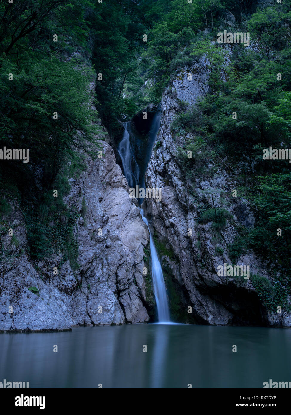 A tall waterfall falls from a cliff into a clear lake at night, lit by ...